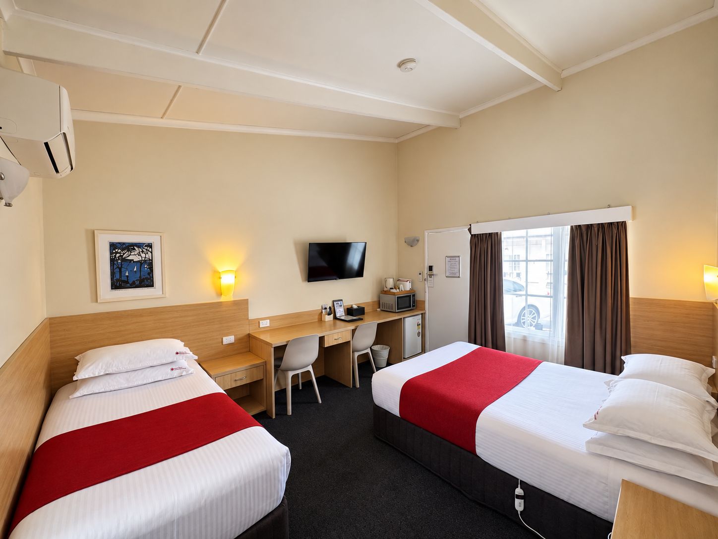 Quarto de hotel com duas camas, uma escrivaninha, televisão e janela com cortinas.