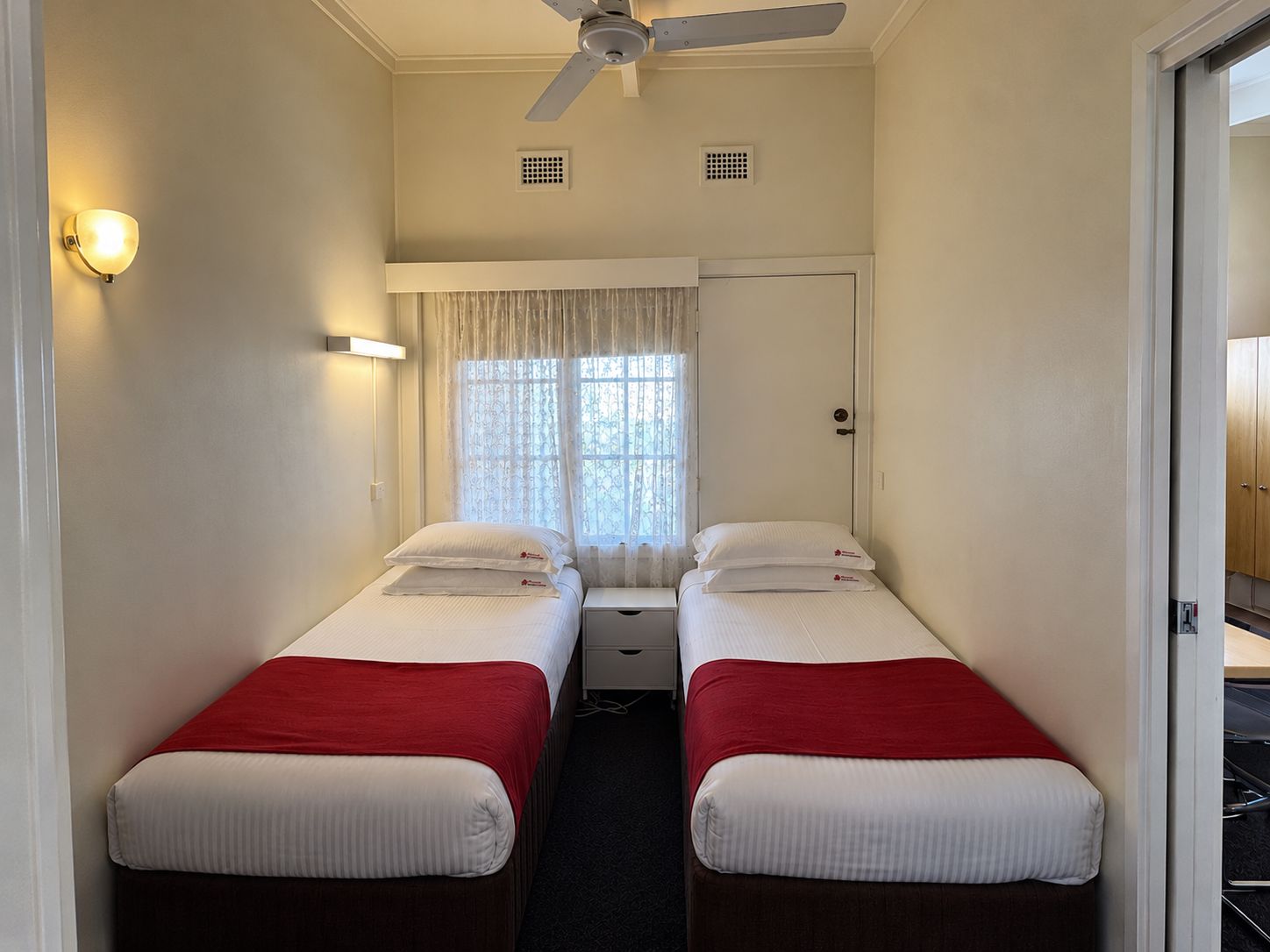 Quarto de hotel com duas camas, um ventilador de teto, uma janela com cortinas e uma porta que dá para uma varanda.