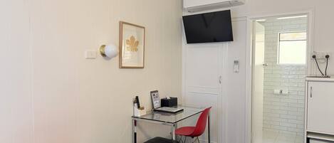 Quarto compacto com escrivaninha de vidro, cadeira vermelha, quadro emoldurado, televisão preta e ar condicionado branco.