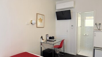 Quarto compacto com escrivaninha de vidro, cadeira vermelha, quadro emoldurado, televisão preta e ar condicionado branco.