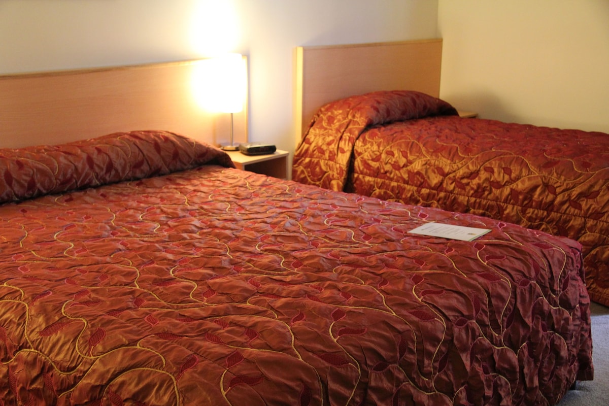 Quarto de hotel com duas camas, cada uma com colcha vermelha estampada e uma mesa de cabeceira com abajur.