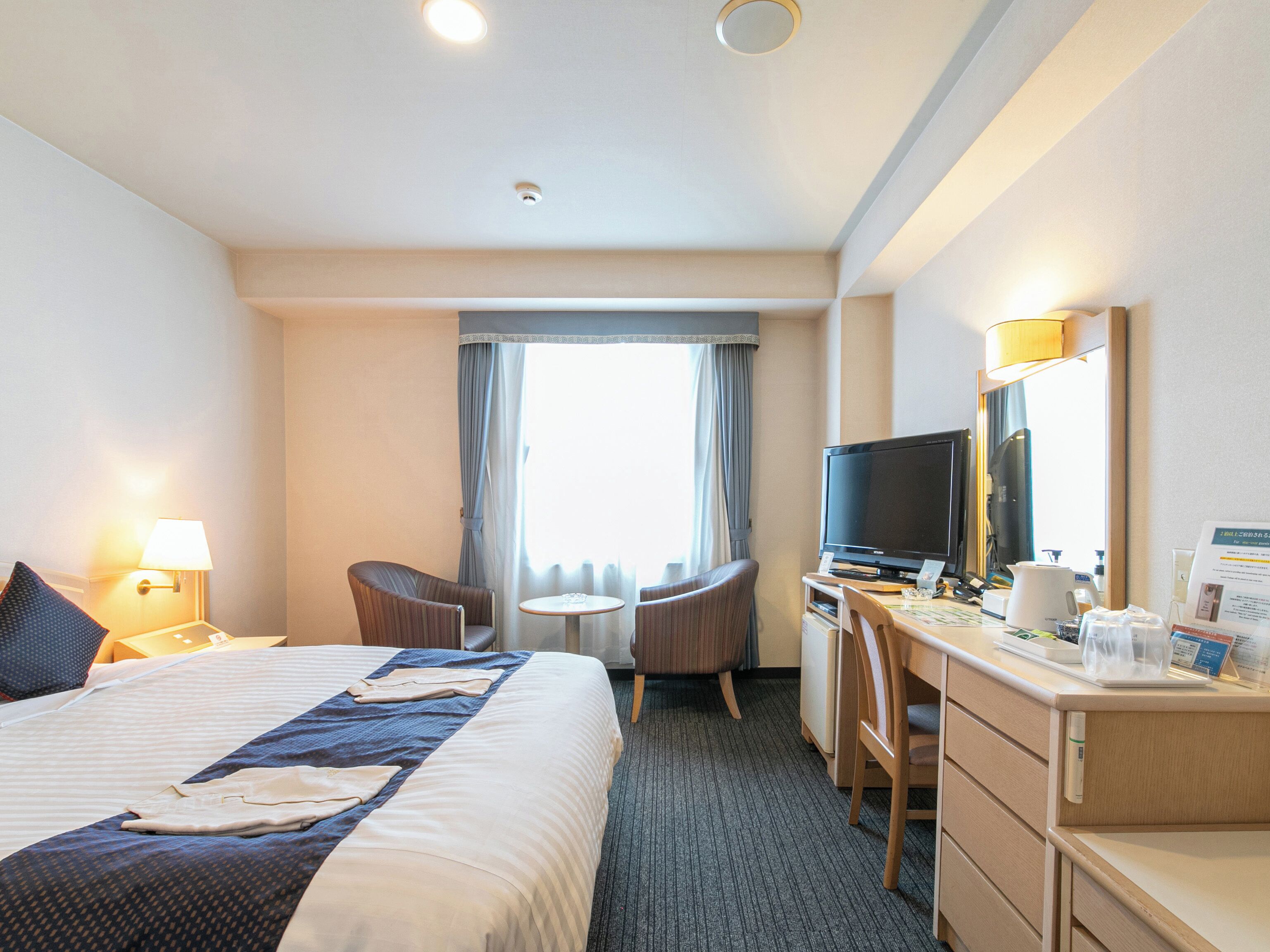 Quarto de hotel com uma cama, uma escrivaninha com computador, uma cadeira, televisão e janela com cortinas.