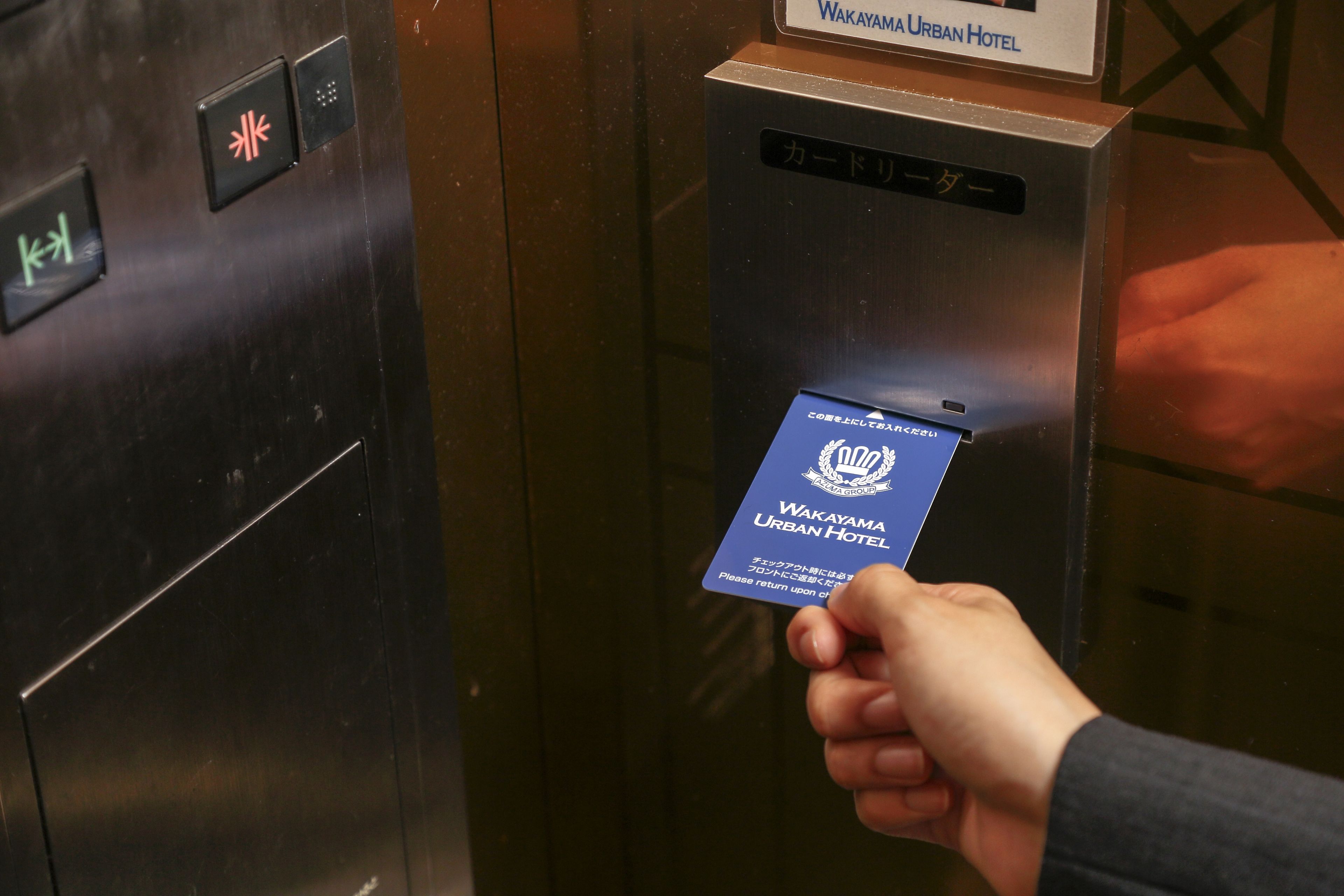 Uma mão segurando um cartão com o texto 'Wakayama Urban Hotel' em frente a um elevador com uma placa acima.