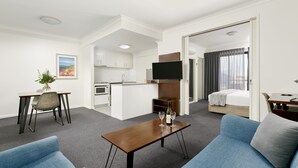 Quarto de hotel com cozinha compacta, área de refeições, sala de estar com sofá e quarto com cama e cortinas.