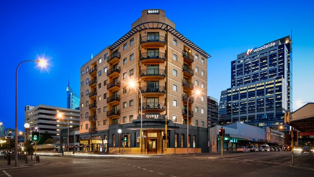 Um hotel de vários andares com varandas, um letreiro chamativo com a palavra 'QUEST' e um edifício com estilo rústico ao fundo.