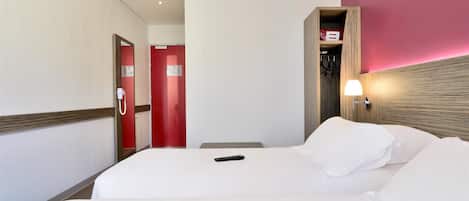 Quarto de hotel com uma cama, mesa de cabeceira com abajur, porta vermelha e cabeceira de madeira.