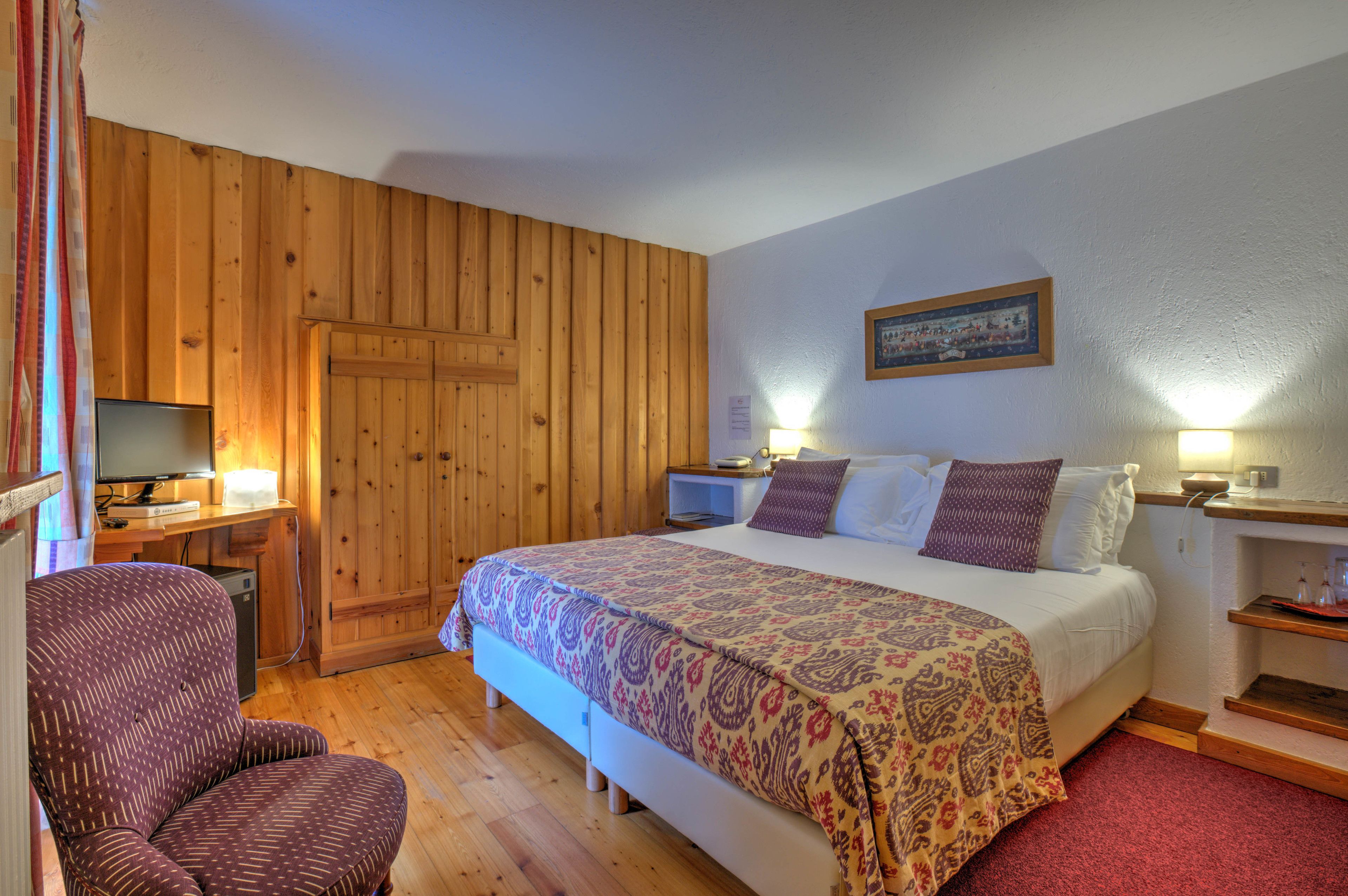 Quarto com painel de parede em madeira, cama com roupa de cama estampada, uma cadeira, escrivaninha com computador e mesa de cabeceira com abajur.