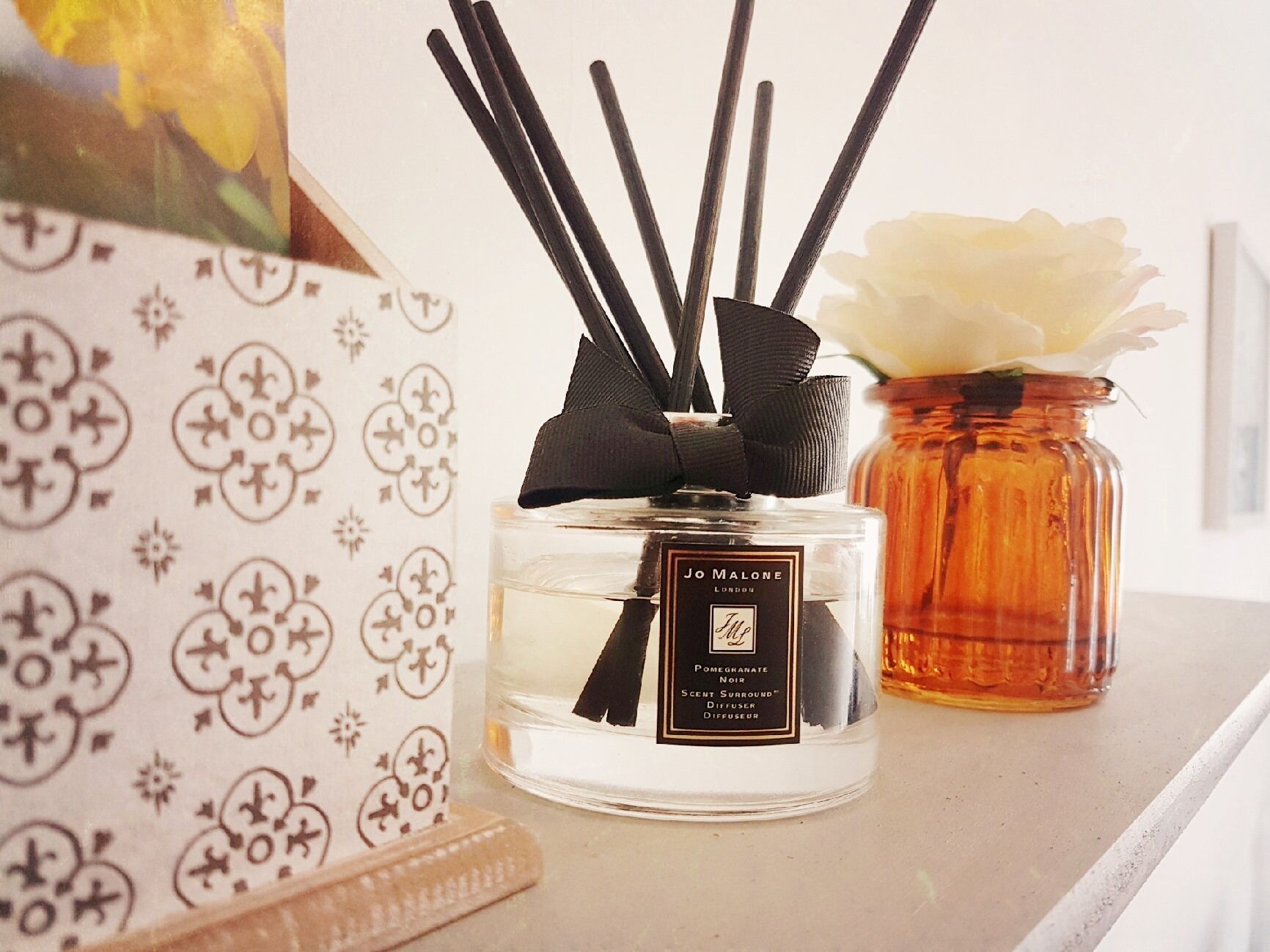 Um difusor de fragrância Jo Malone London Pomegranate Noir, em garrafa de vidro transparente com canudos pretos, colocado sobre uma mesa com papel estampado e um vaso com flores brancas.