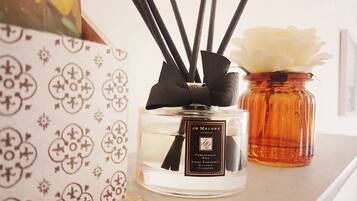 Um difusor de fragrância Jo Malone London Pomegranate Noir, em garrafa de vidro transparente com canudos pretos, colocado sobre uma mesa com papel estampado e um vaso com flores brancas.