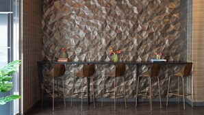 Uma área de bar moderna com parede estampada em padrão geométrico, dois bancos e uma pequena mesa com um vaso de flores.