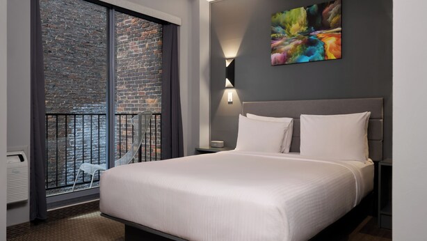 Quarto de hotel moderno com uma cama grande, vista para uma parede de tijolos, varanda com uma cadeira e uma pintura abstrata colorida na parede.