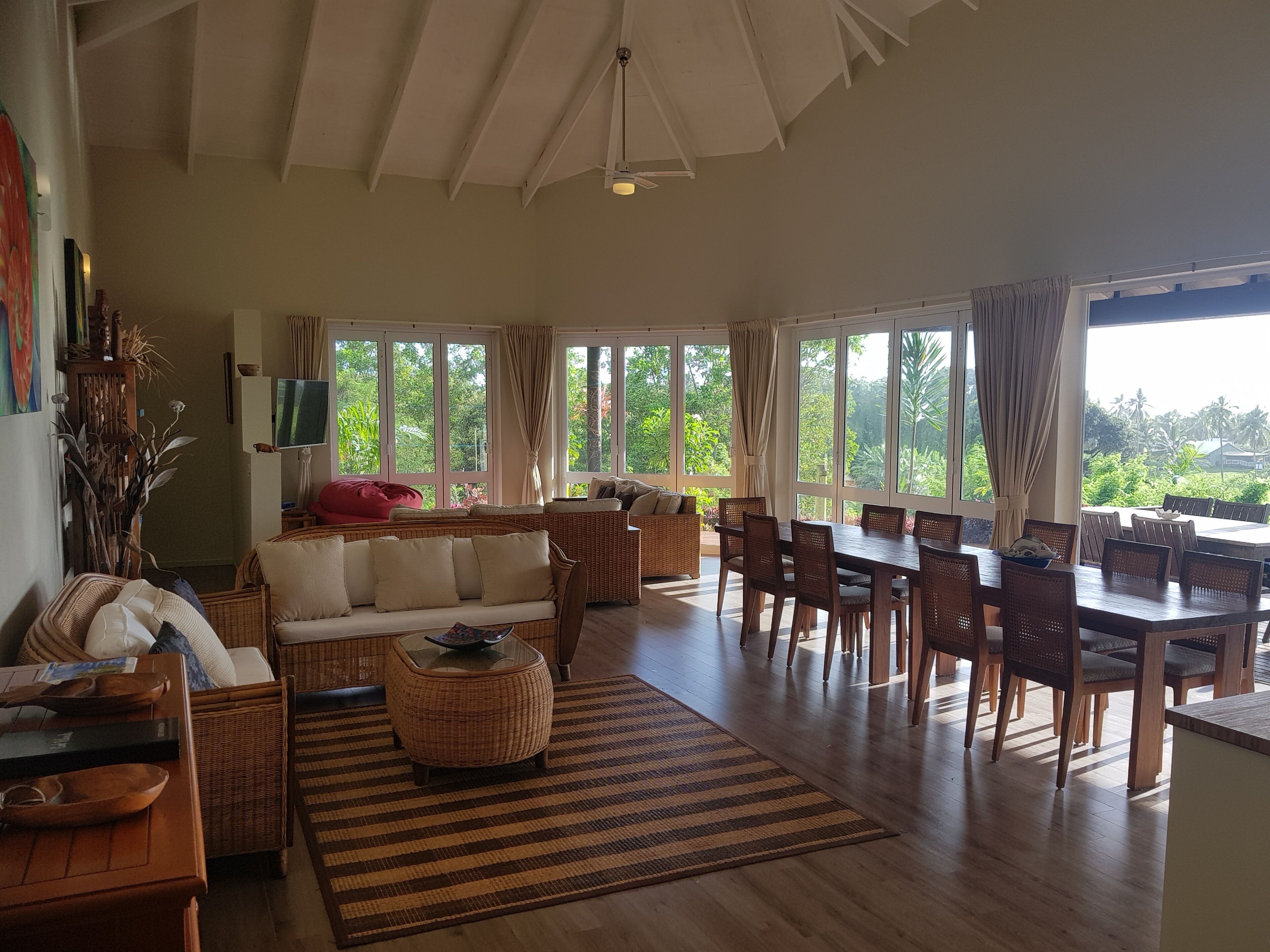 Uma sala de estar espaçosa com sofá, mesa de jantar e cadeiras, janelas amplas com cortinas e um ventilador de teto.