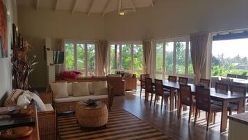 Uma sala de estar espaçosa com sofá, mesa de jantar e cadeiras, janelas amplas com cortinas e um ventilador de teto.