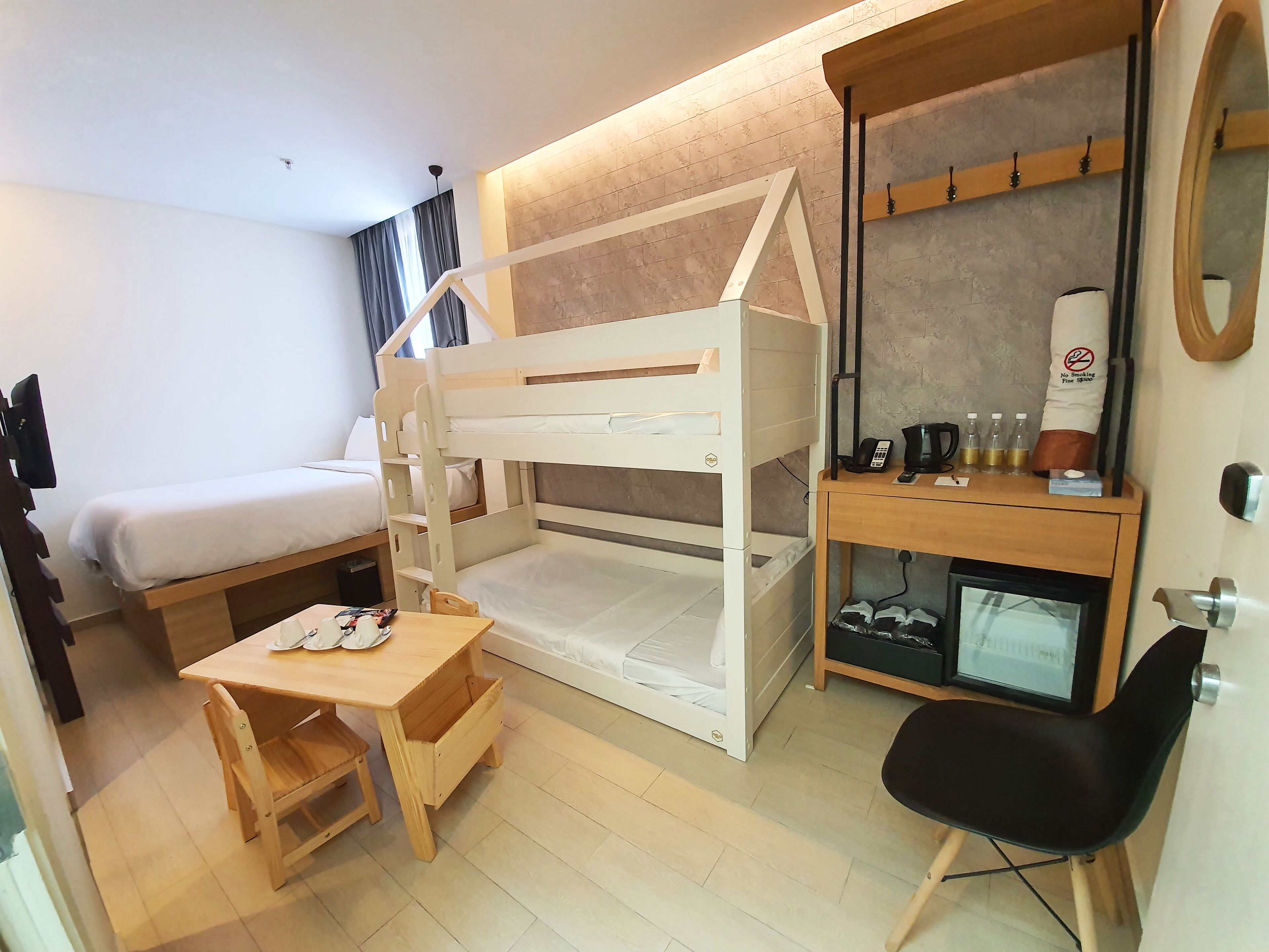 Quarto de hotel pequeno com uma cama beliche, uma escrivaninha de madeira, uma cadeira, uma mesa de cabeceira, uma televisão e um espelho.
