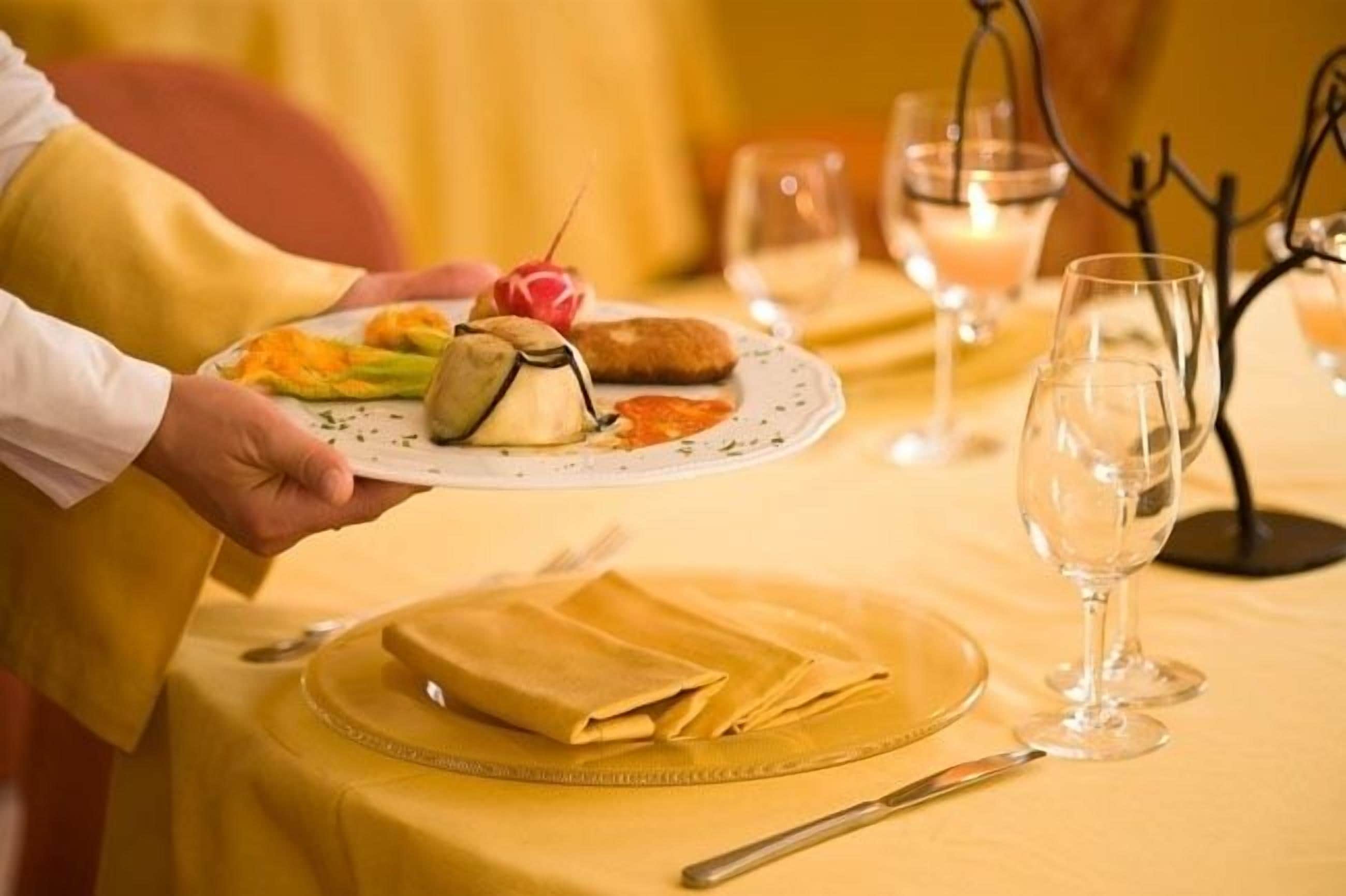 Uma pessoa servindo um prato de comida a uma mesa posta com toalha amarela, copos de vinho e castiçal.