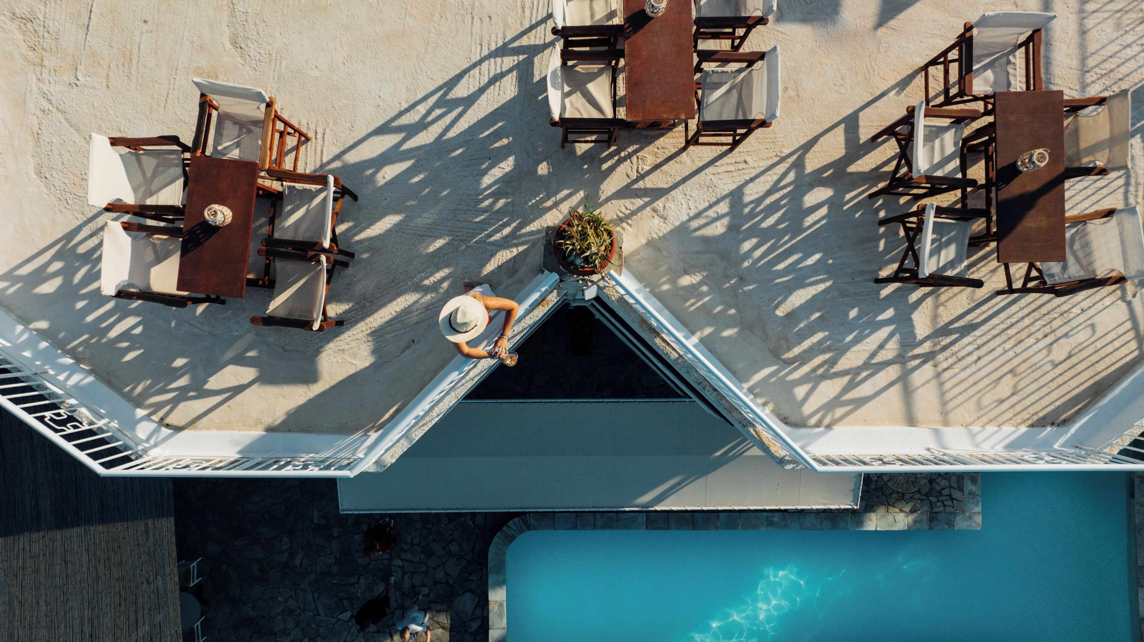 Vista aérea de uma área de piscina no terraço, com várias cadeiras de sol e mesas, um vaso de planta e uma pessoa sentada em uma cadeira.