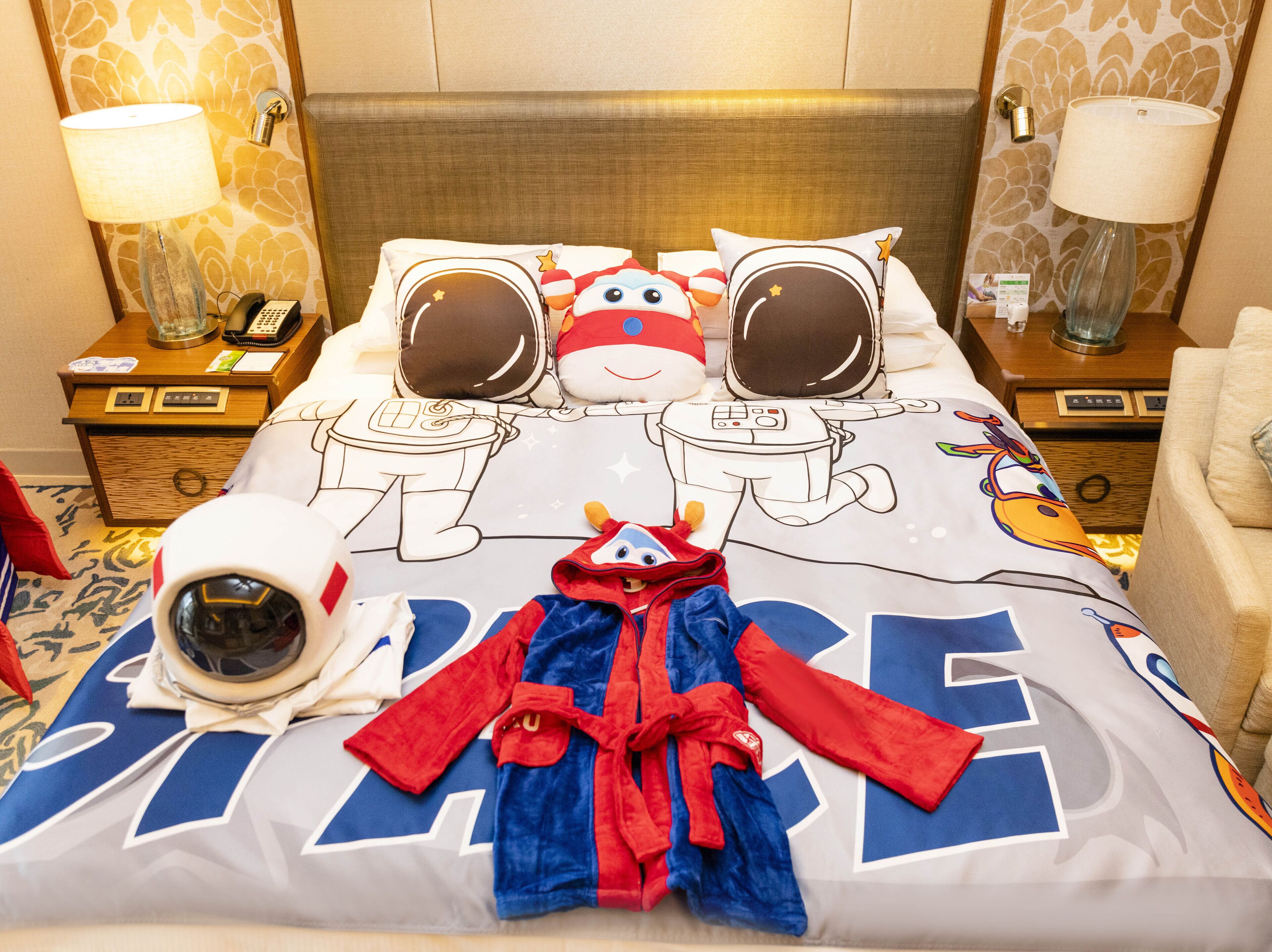 Quarto de hotel com uma cama, um edredom com tema espacial, um brinquedo de pelúcia e um capacete espacial sobre a cama.
