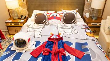 Quarto de hotel com uma cama, um edredom com tema espacial, um brinquedo de pelúcia e um capacete espacial sobre a cama.