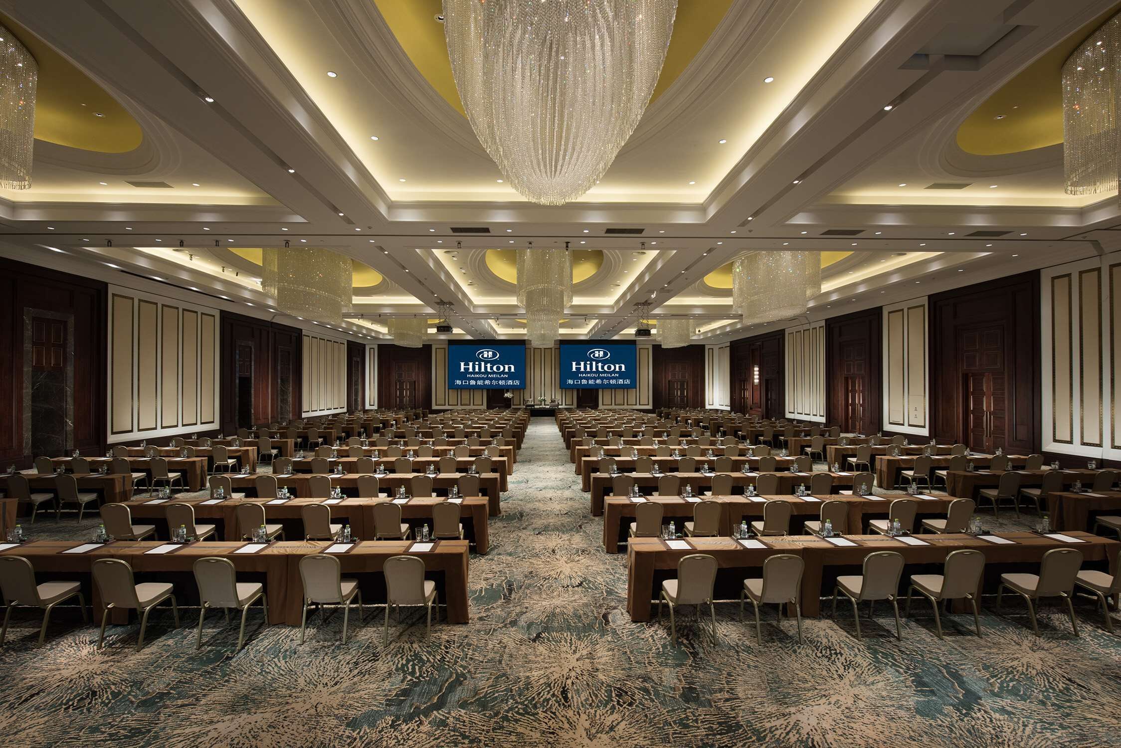 Uma sala de conferências espaçosa com fileiras de mesas e cadeiras, lustres e sinalização Hilton.