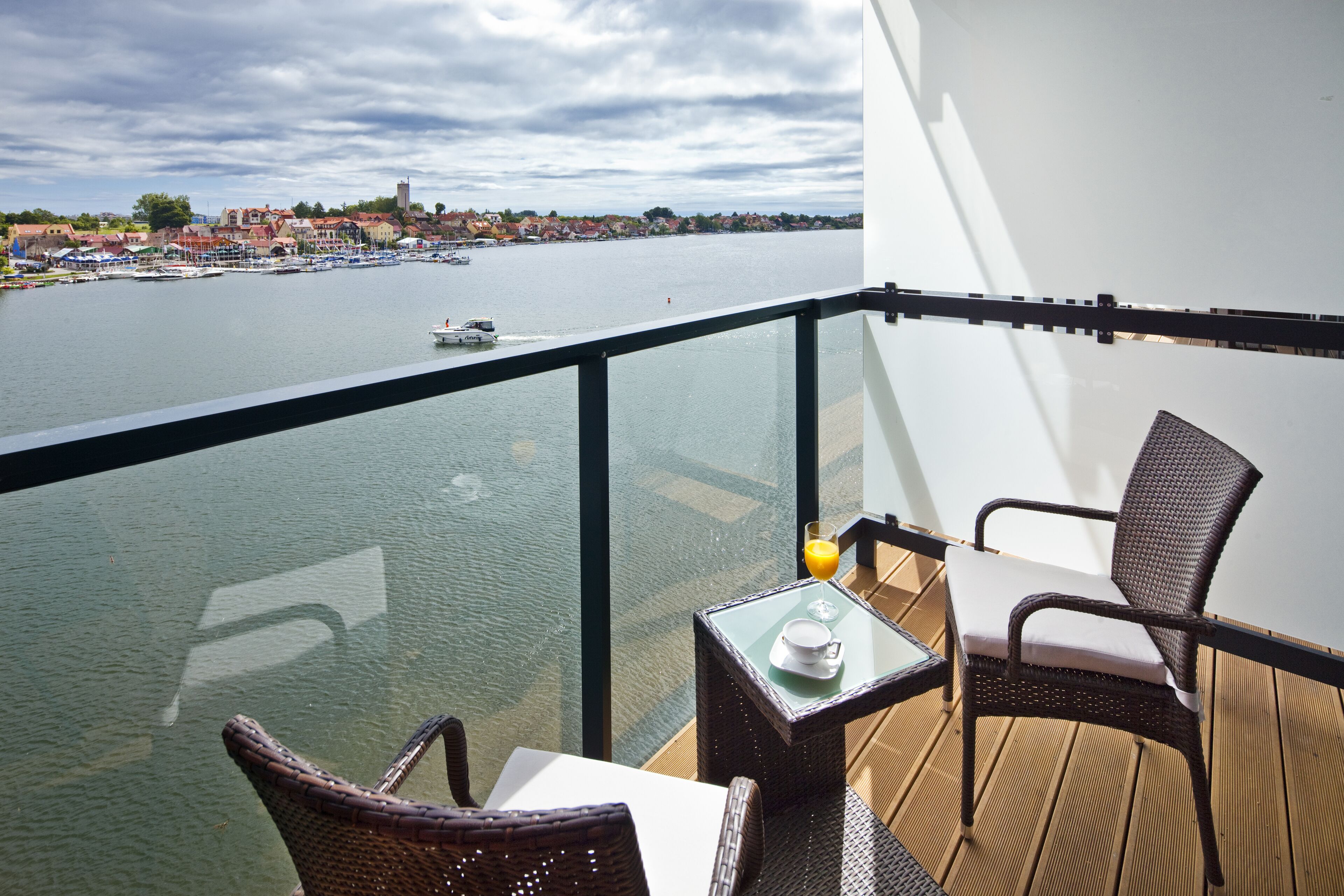 Uma varanda com móveis de vime, uma mesa de vidro e vista para um rio com barcos e edifícios ao longe.