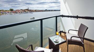 Uma varanda com móveis de vime, uma mesa de vidro e vista para um rio com barcos e edifícios ao longe.