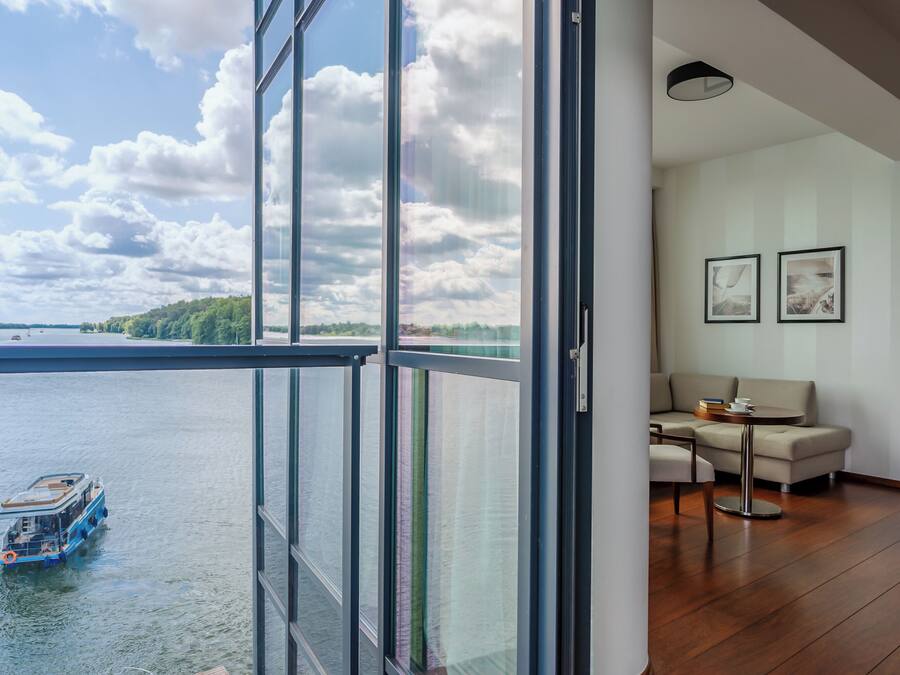 Quarto com uma grande janela com vista para um lago, um sofá, uma mesa pequena e duas fotografias emolduradas na parede.