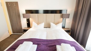 Cama de casal com lençóis brancos, manta roxa e duas toalhas brancas dobradas ao pé da cama. Há duas mesas de cabeceira com luminárias, uma porta de madeira e uma cabeceira com luminária.