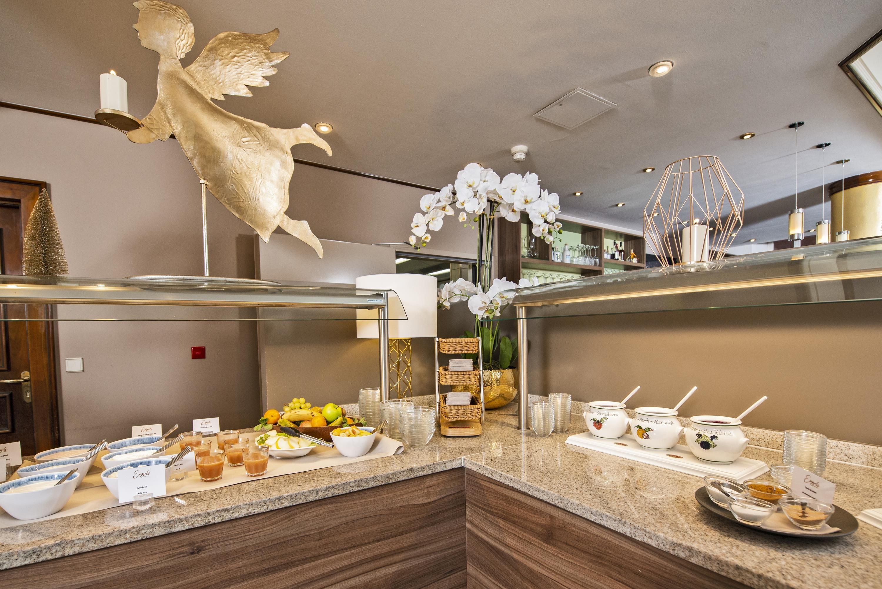 Uma área de buffet com diversas opções de comida, incluindo frutas, doces e bebidas, dispostas em um balcão com uma escultura decorativa de anjo acima.