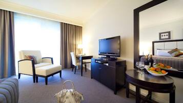Quarto de hotel com sofá, poltrona, televisão, mesa de jantar com frutas e bebidas, e cama com cabeceira.