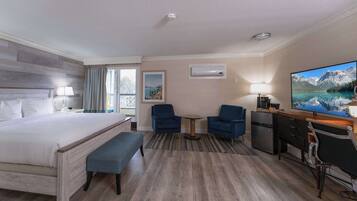 Quarto de hotel com uma cama grande, uma escrivaninha com computador, duas poltronas, uma mesa pequena e uma televisão exibindo uma paisagem de montanhas.
