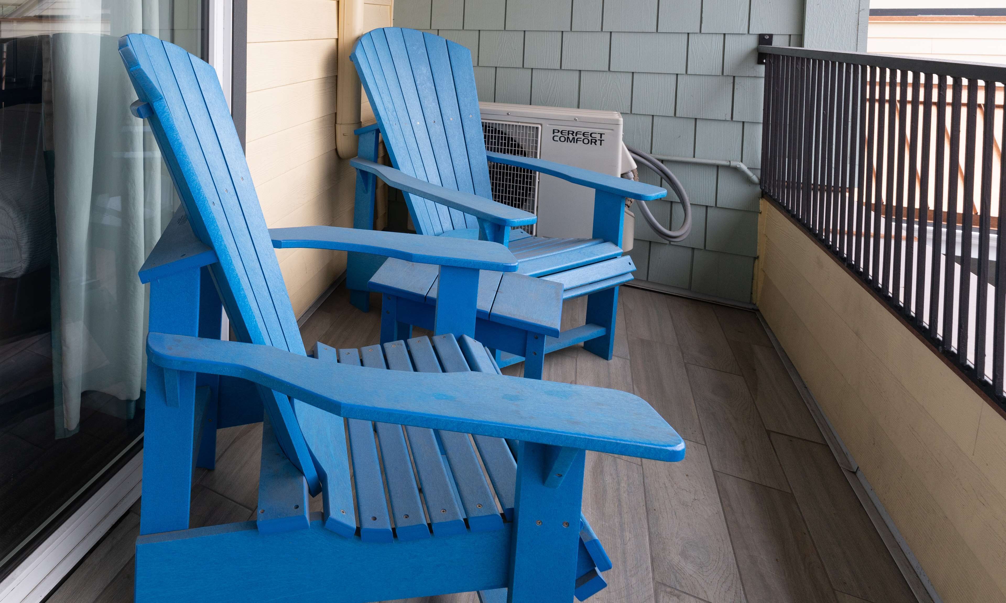 Duas cadeiras Adirondack azuis sobre um deck de madeira com guarda-corpo metálico e uma janela com cortina branca.