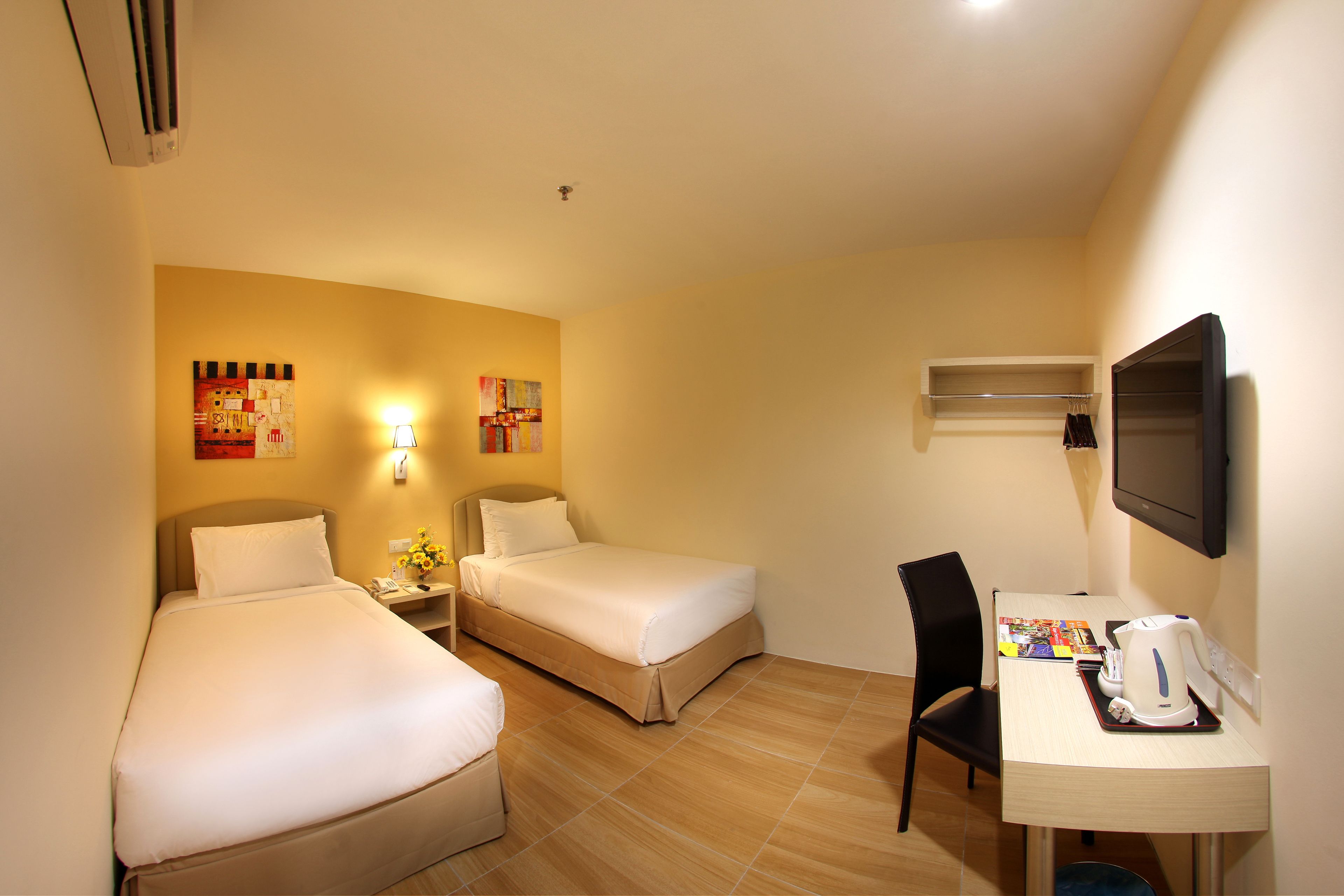 Quarto de hotel com duas camas, uma escrivaninha com chaleira, uma cadeira e uma TV de tela plana na parede.