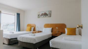 Quarto de hotel com três camas de solteiro, cada uma com cabeceira de madeira, roupa de cama branca e travesseiro amarelo. Há uma janela com cortinas azuis, uma obra de arte na parede e uma pequena mesa com luminária.