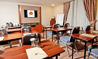 Uma sala de conferências de hotel com mesas e cadeiras de madeira, um quadro branco, uma tela de TV e um dispensador de água.