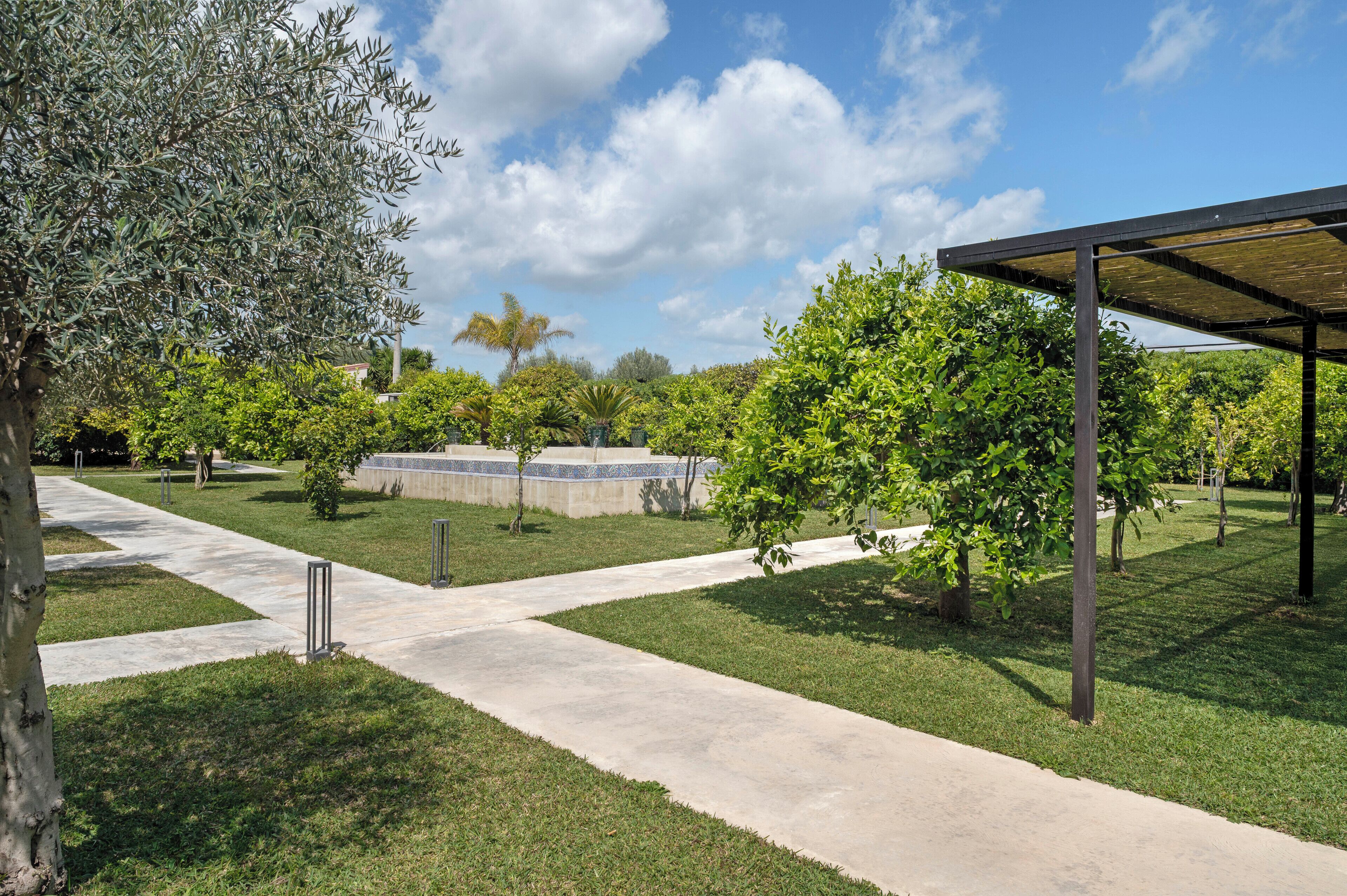 Um jardim ajardinado com um caminho pavimentado, uma pérgola e diversas árvores.
