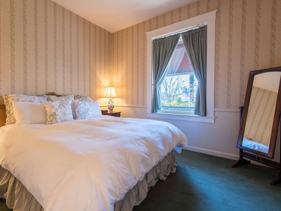 Cama bem arrumada com cabeceira estampada em flor, luminária de cabeceira e janela com cortinas.
