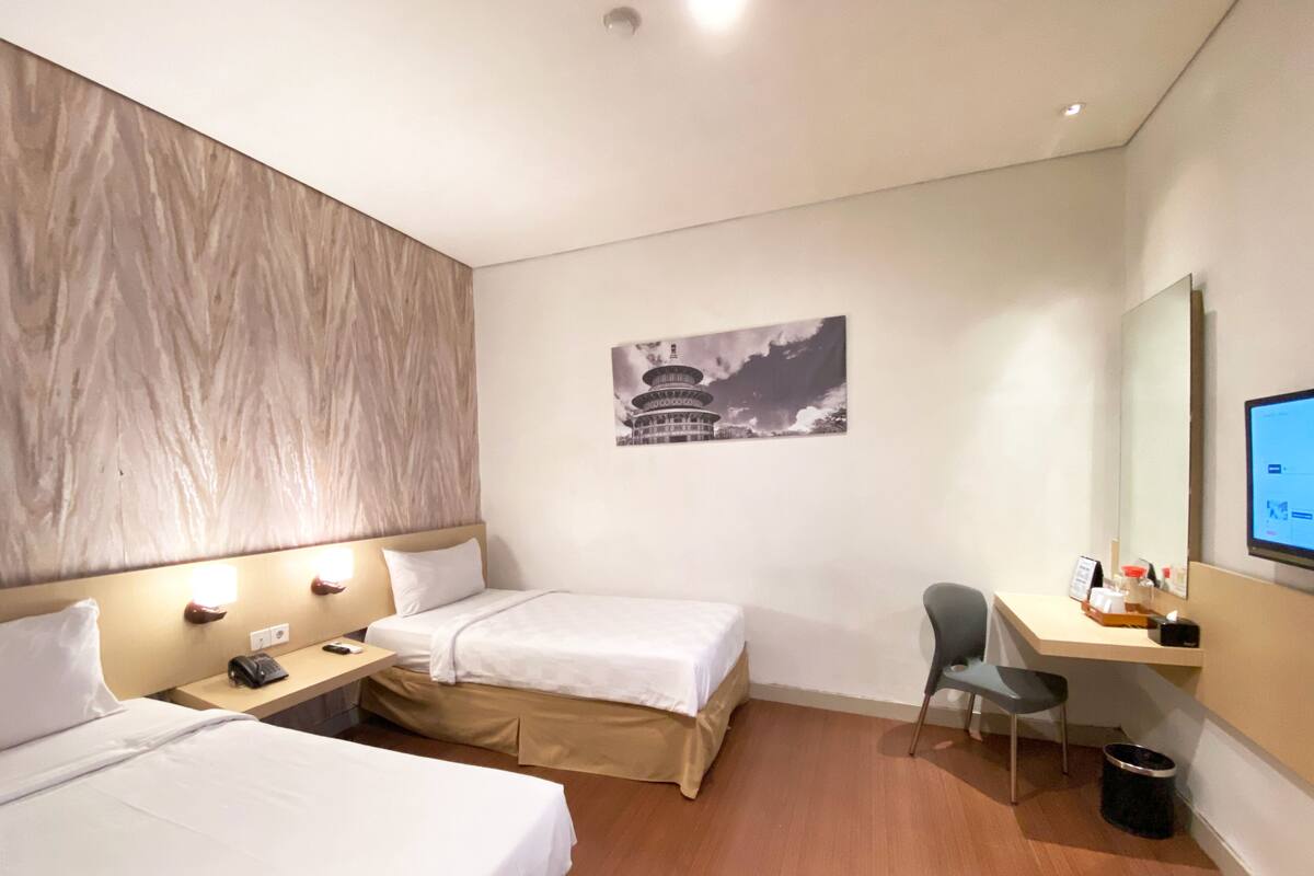 Quarto de hotel com duas camas, uma escrivaninha com cadeira, um cesto de lixo pequeno e uma TV fixada na parede.