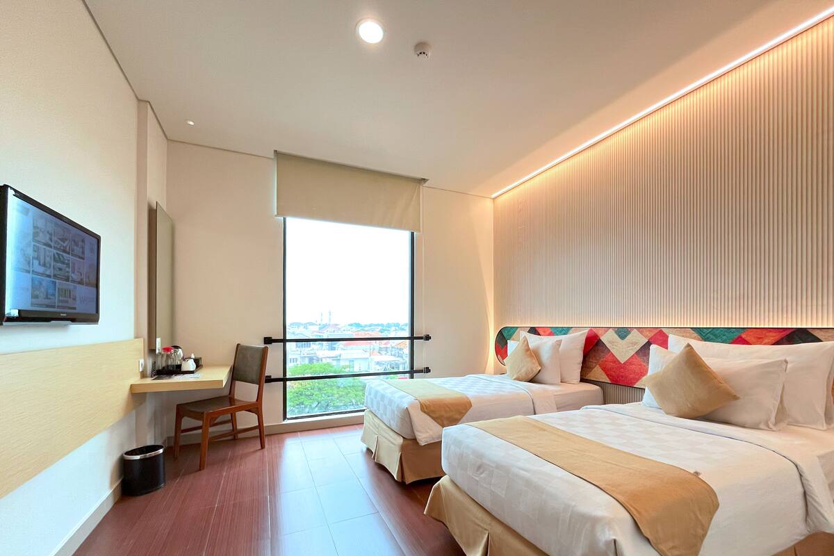 Quarto de hotel com duas camas, uma escrivaninha, uma cadeira, uma TV e uma janela grande com vista para a cidade.