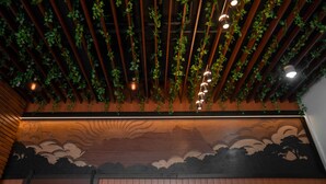 Teto com vigas de madeira verticais e trepadeiras verdes pendentes, iluminado por vários luminários.