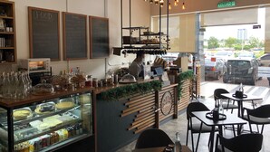 Um café com um balcão exibindo comida e bebidas, uma máquina de café e uma área de estar com mesas e cadeiras.