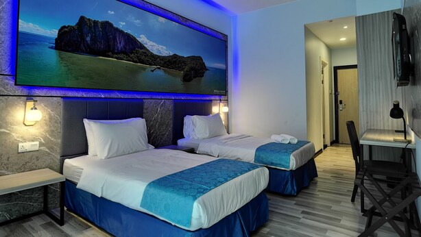 Quarto de hotel com duas camas, uma televisão grande fixada na parede exibindo uma paisagem, uma escrivaninha com cadeira e uma porta que dá para outro cômodo.