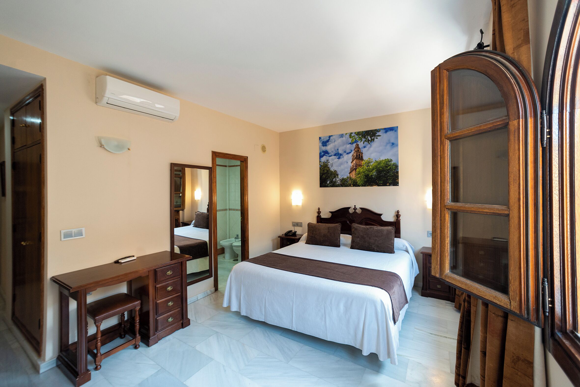 Quarto de hotel com cama, escrivaninha, guarda-roupa, espelho e um quadro na parede.