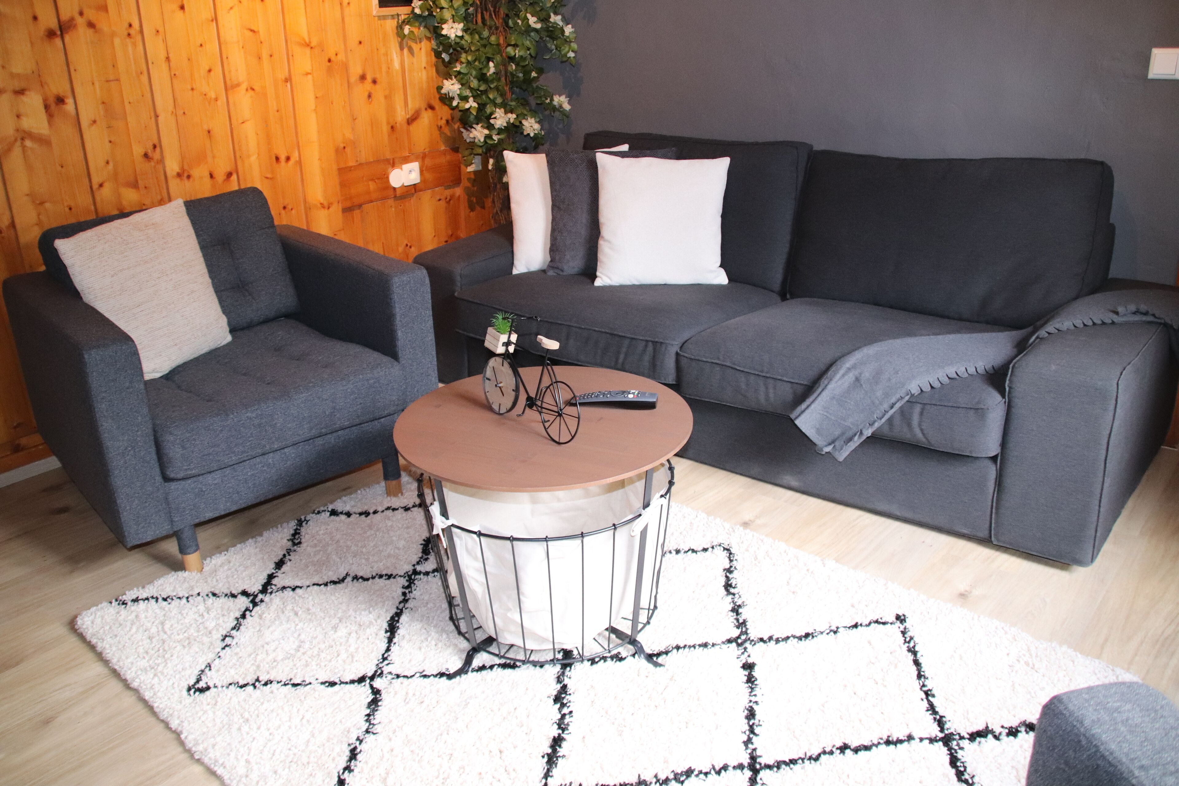 Uma sala de estar com um sofá cinzento, uma poltrona cinza menor, uma mesa de centro redonda com uma planta e um modelo de bicicleta, e um tapete estampado.