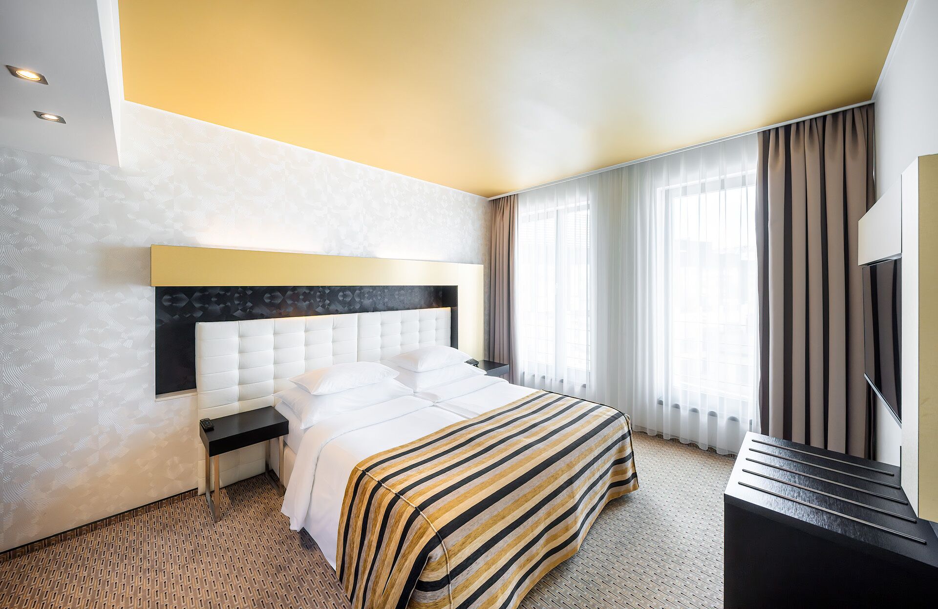Quarto de hotel com uma cama grande, mesa de cabeceira, televisão e janela com cortinas.