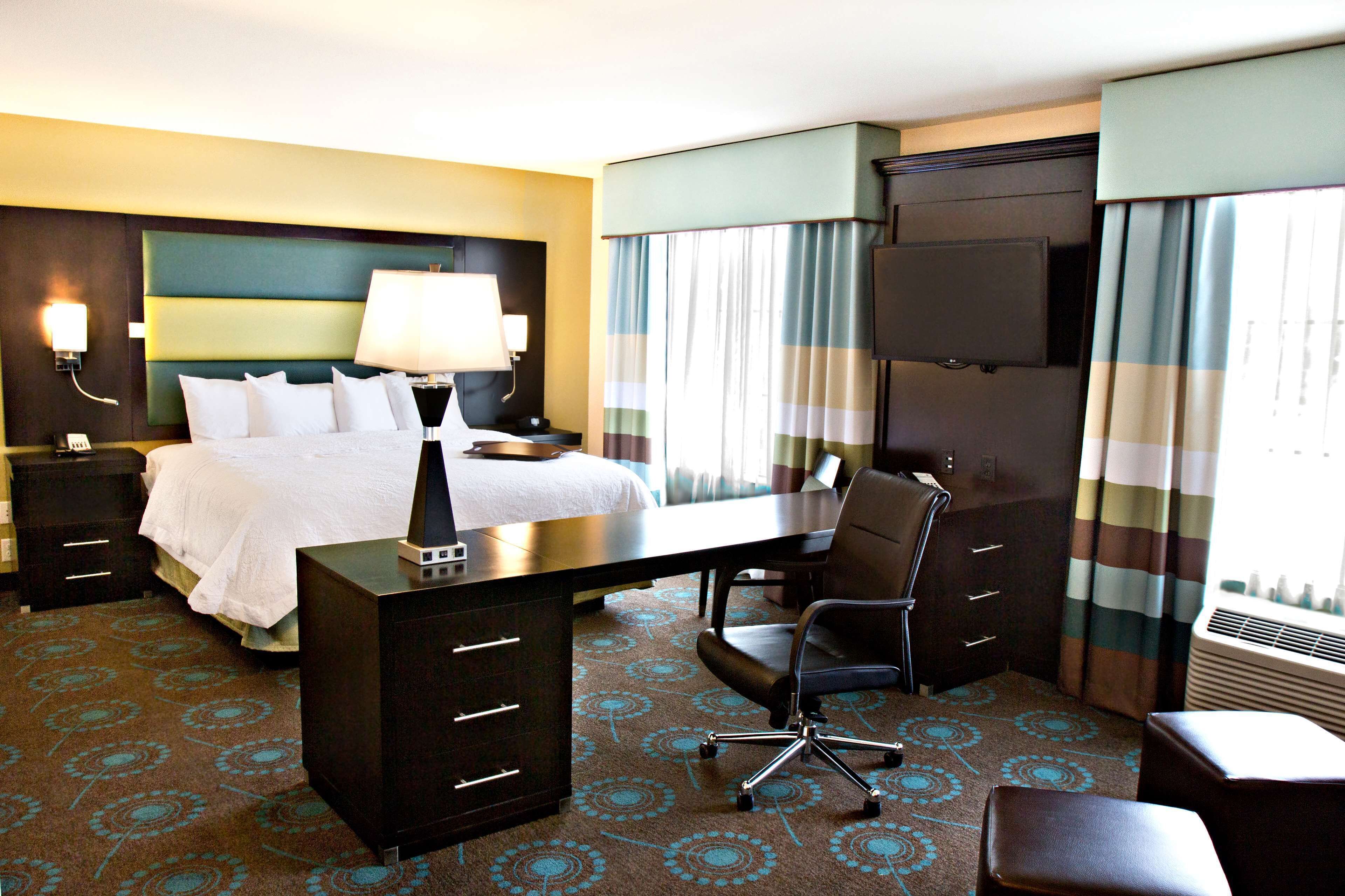 Quarto de hotel com cama, escrivaninha, cadeira e televisão.