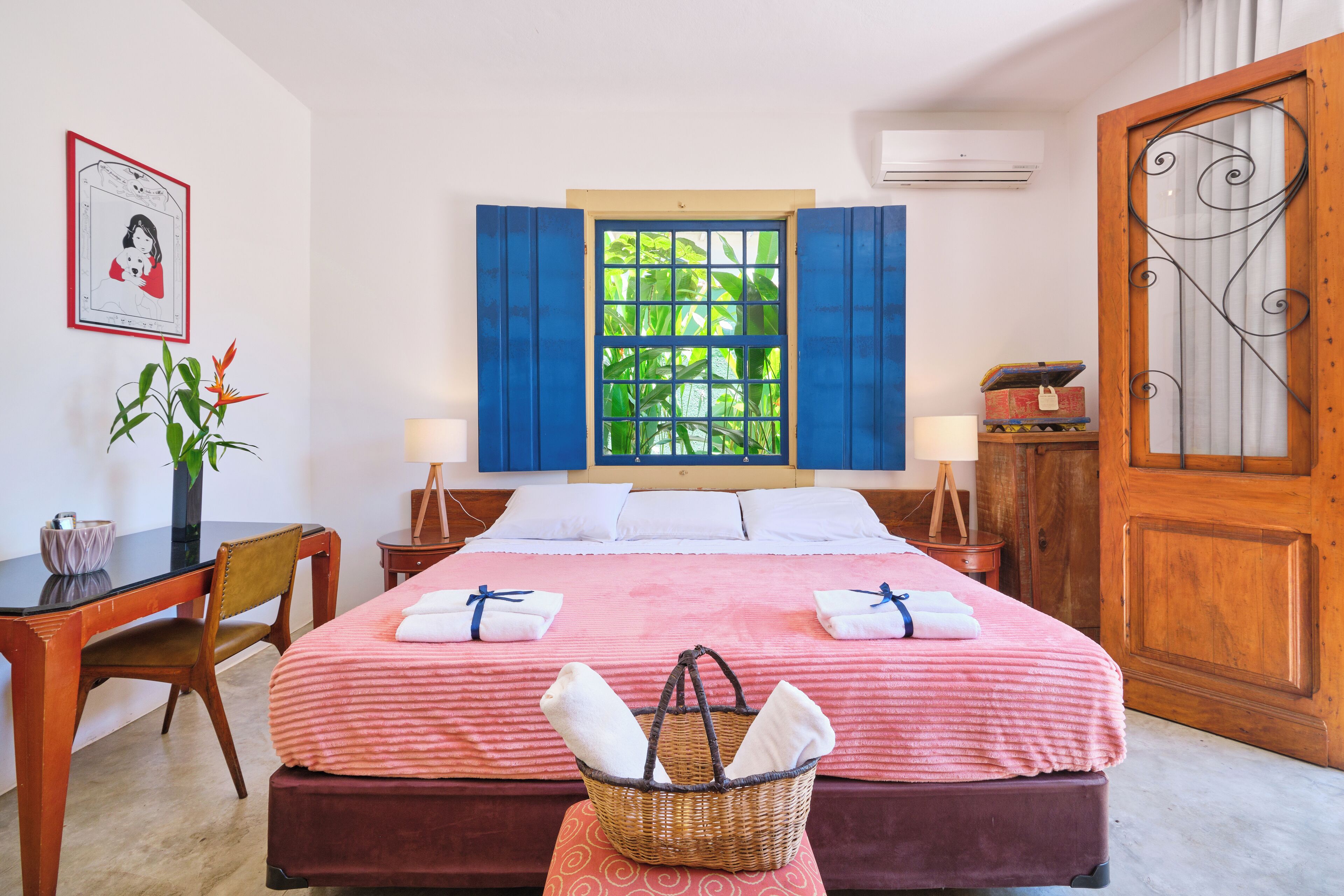 Quarto de hotel com cama, escrivaninha, cadeira, janela com persianas azuis, porta com detalhe em metal e ar-condicionado fixado na parede.