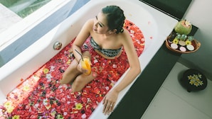 Uma pessoa sentada em uma banheira cheia de pétalas de flores, segurando uma bebida, com um pequeno cesto de produtos de higiene pessoal ao lado.