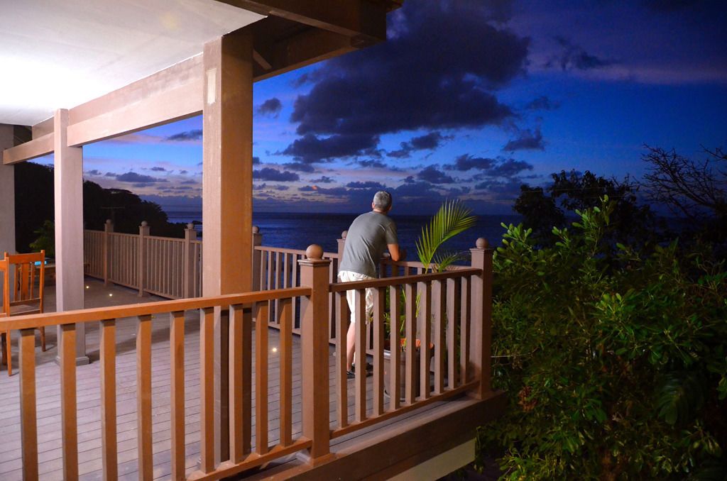 Um homem está em um deck de madeira com guarda-corpo, apreciando a vista cênica do oceano e das colinas ao entardecer.
