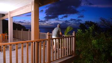 Um homem está em um deck de madeira com guarda-corpo, apreciando a vista cênica do oceano e das colinas ao entardecer.