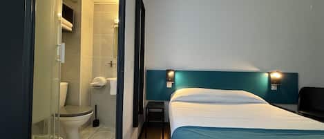 Quarto de hotel com cama, mesa de cabeceira, banheiro com vaso sanitário e pia, e box de vidro.